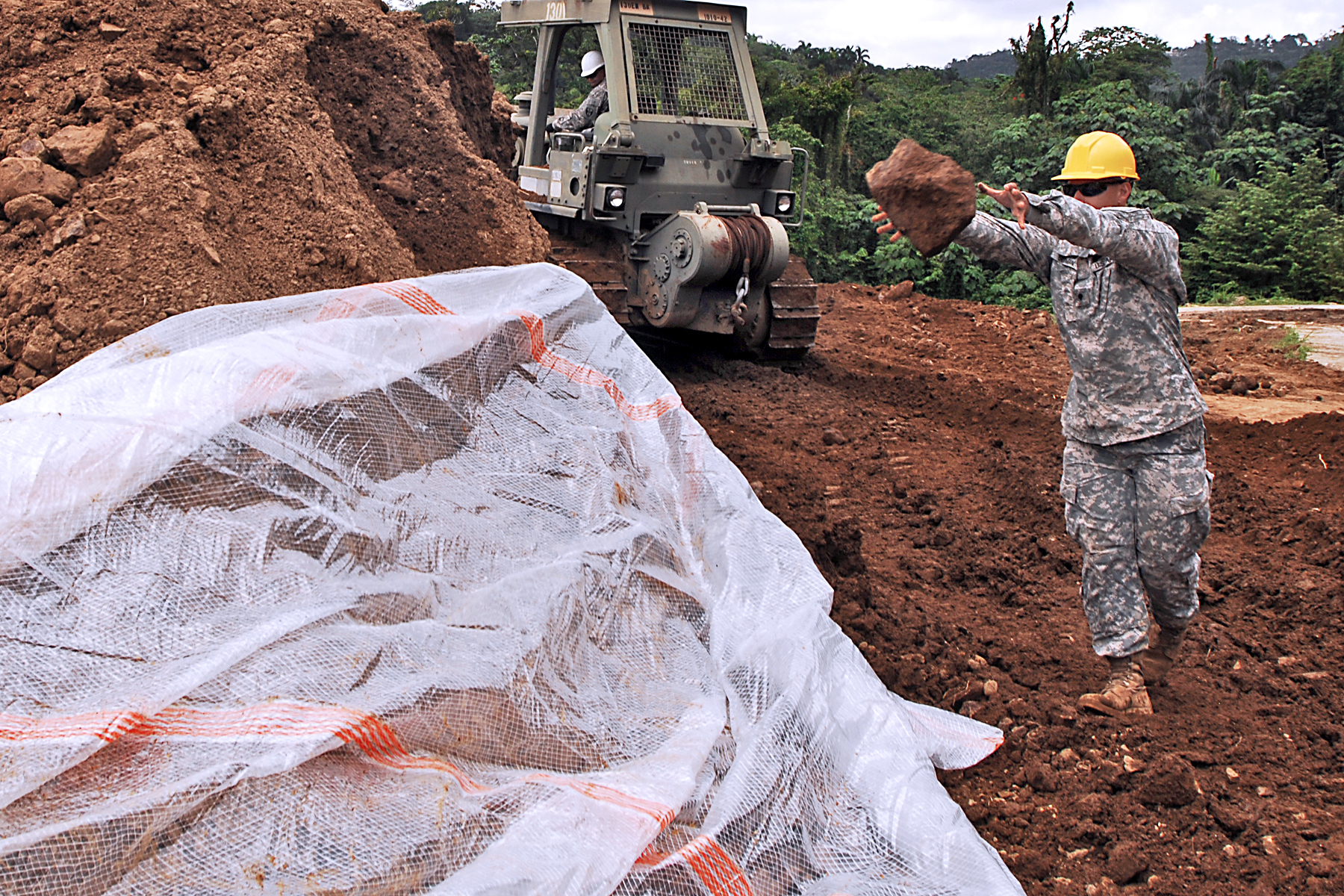 A soldier tosses a rock on top of a plastic tarp that is being used to ...