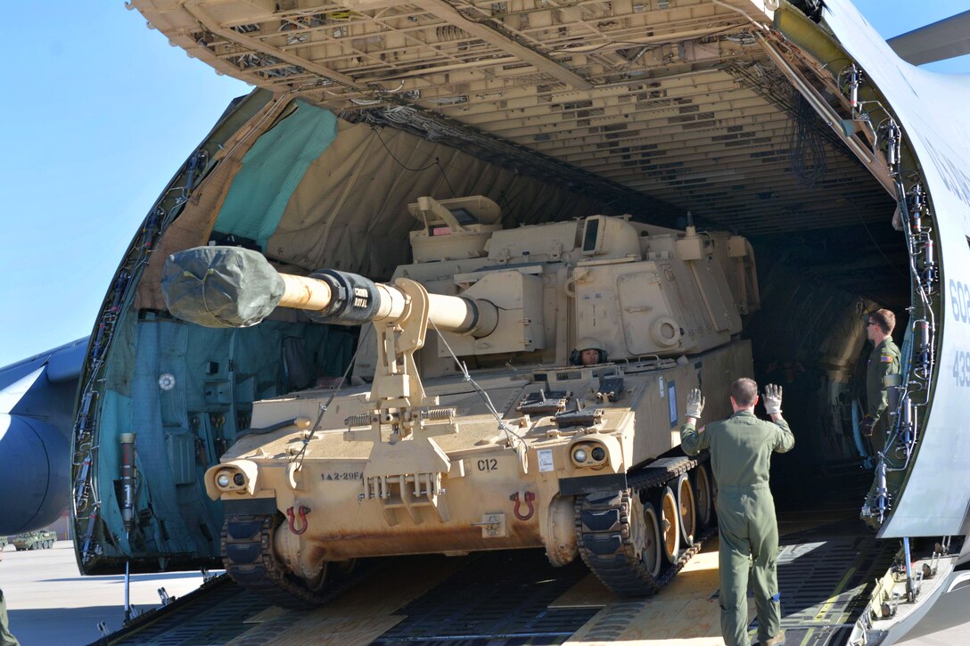 An airman guides a soldier driving a M109A6 Paladin to be loaded onto a ...
